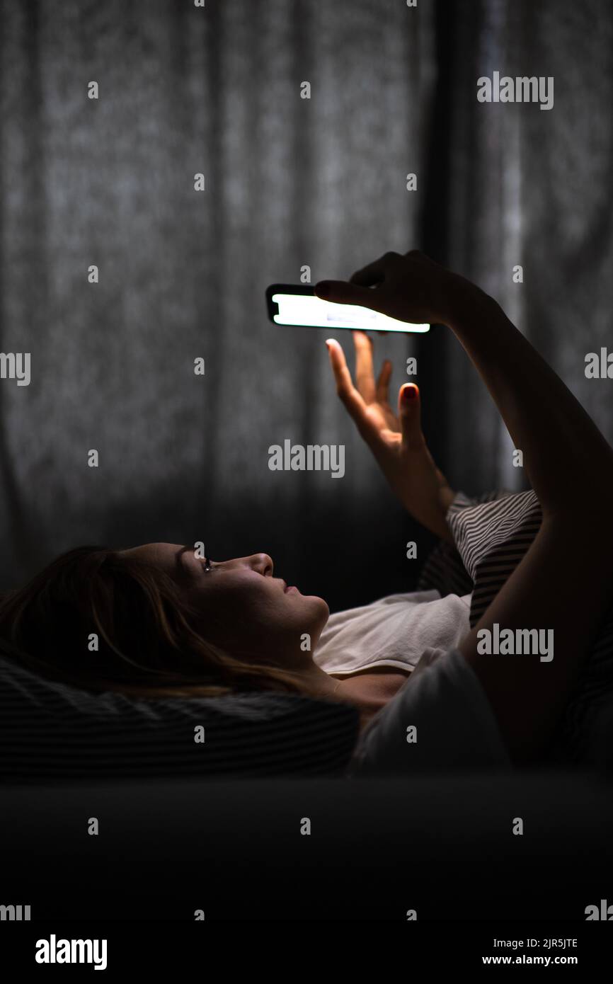 Young woman in bed holding a phone, tired and exhausted, blue light straining her eyes, messing ...