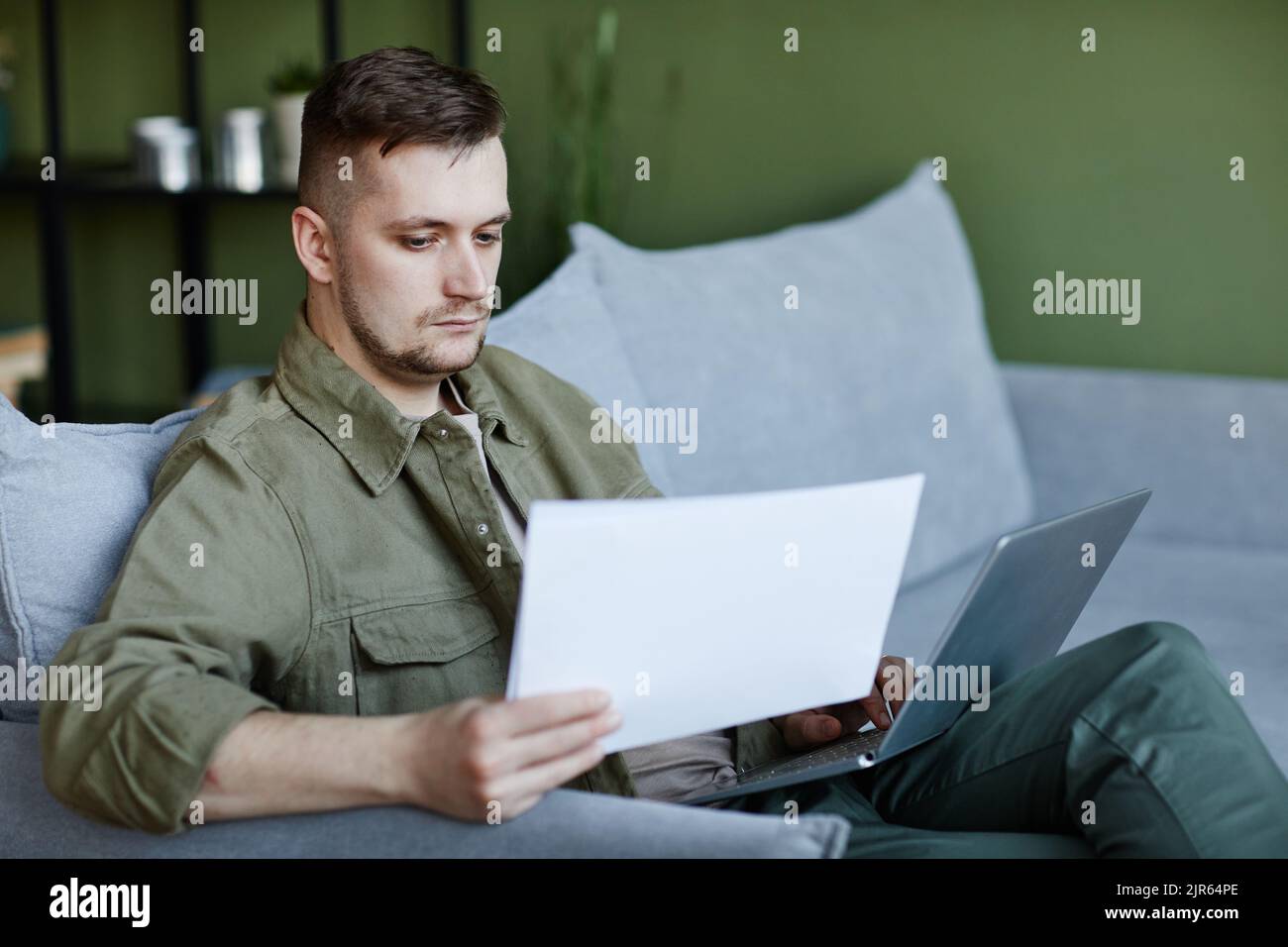 Young serious man sitting on sofa with laptop on his knees,and typing ...