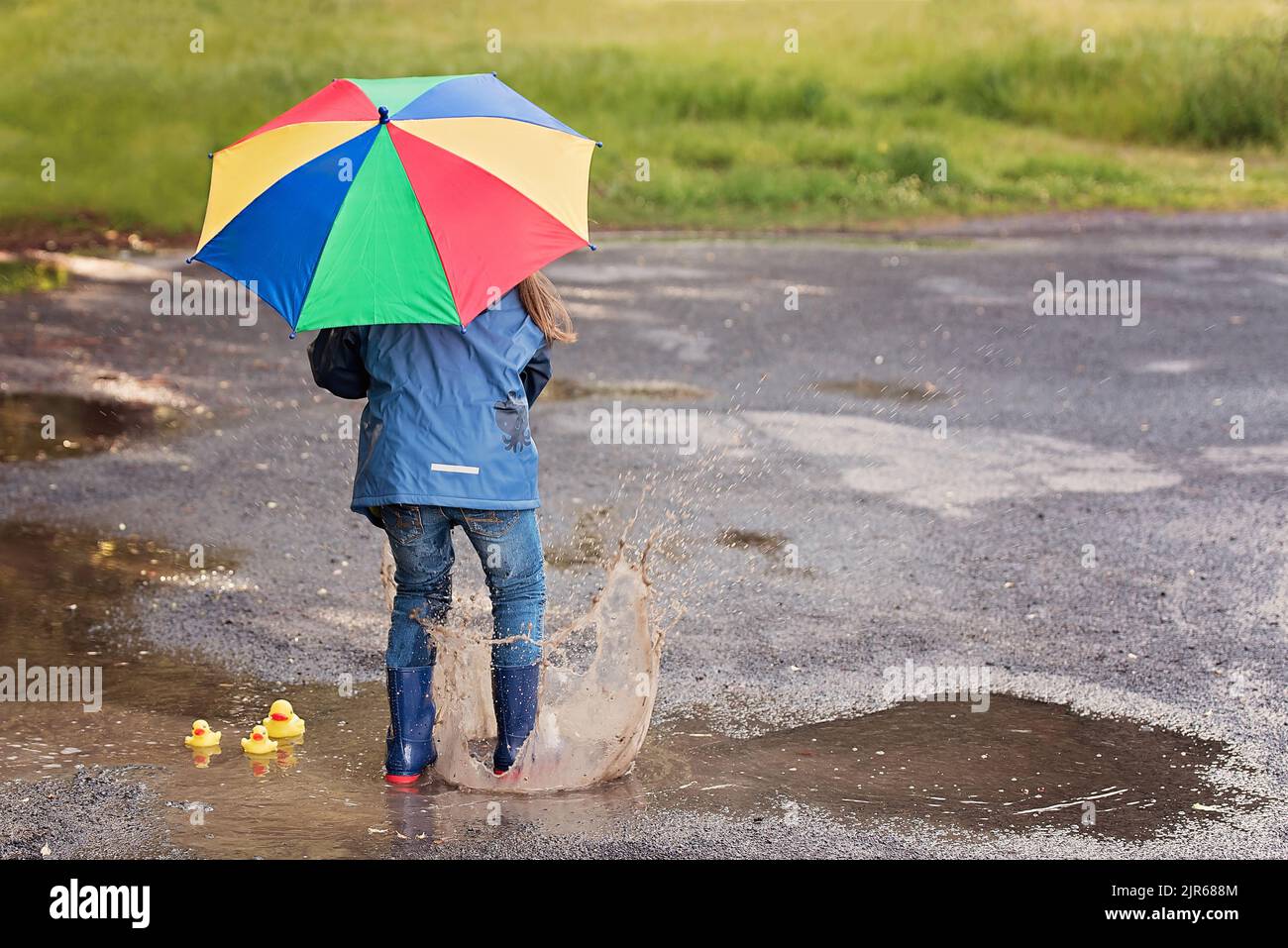 A child holding a colorful umbrella jumping into the rain puddle with ...