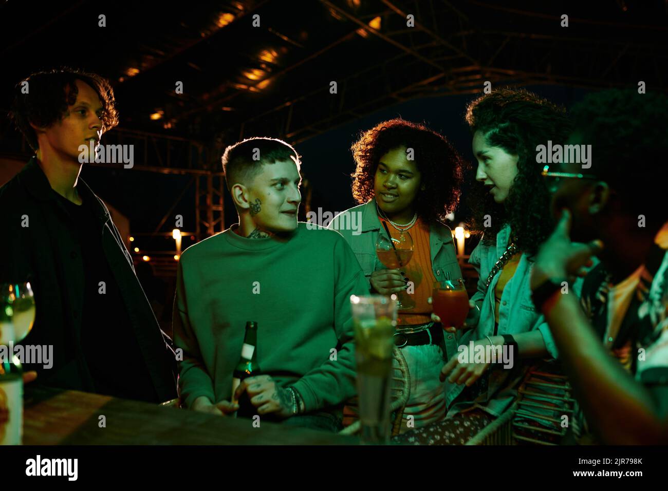 Youthful intercultural girls with cocktails and guys listening to young ...