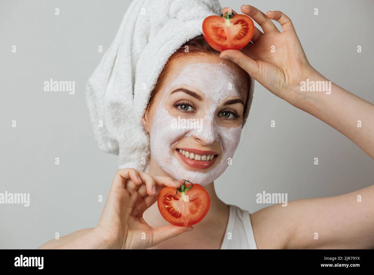 Smiling young woman with white clay mask on face holding fresh tomato ...