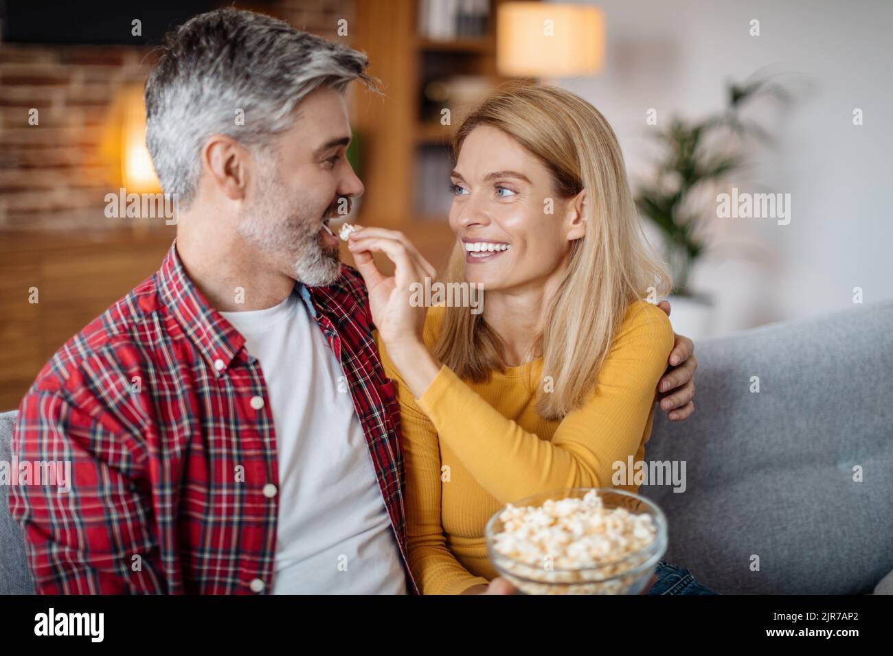 Cheerful middle aged caucasian woman feeding man popcorn, watching tv ...
