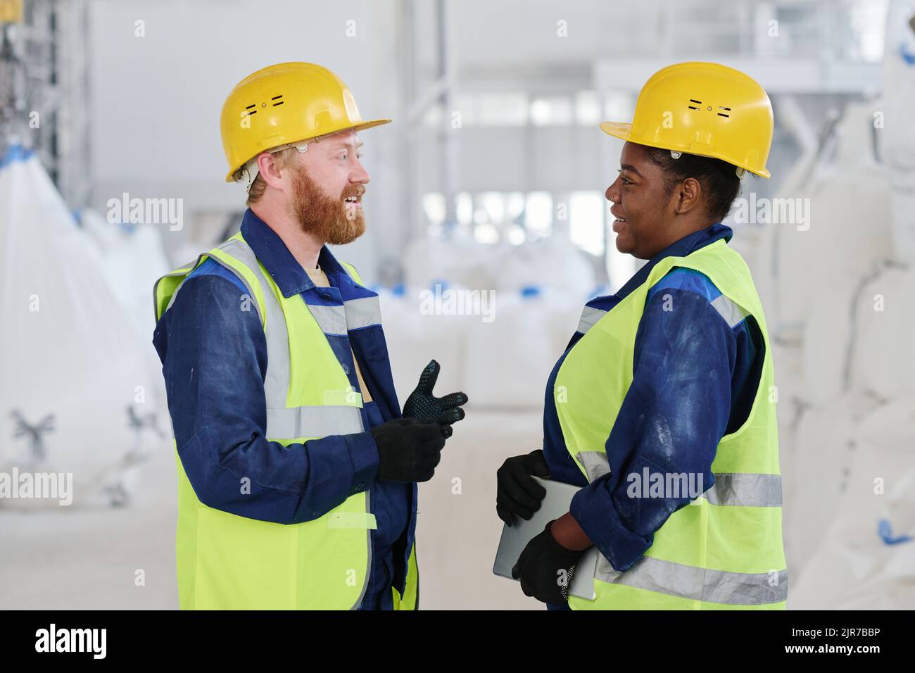 Side view of two young intercultural workers of factory in uniform and hardhats discussing ...