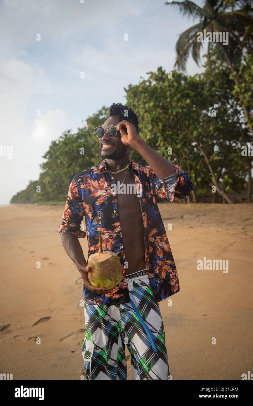 Young man by the sea with a coco nut in hand smiles as he looks at the ...