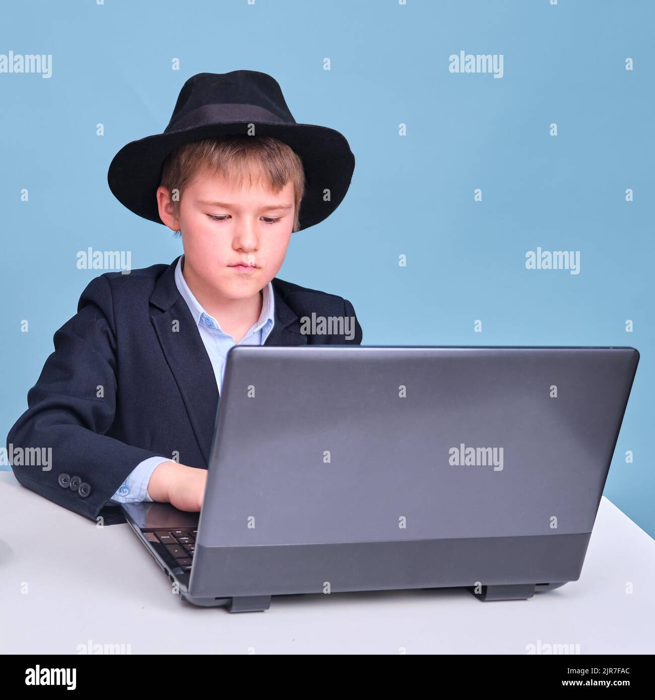 A boy in a school suit and a black hat at a computer during distance ...