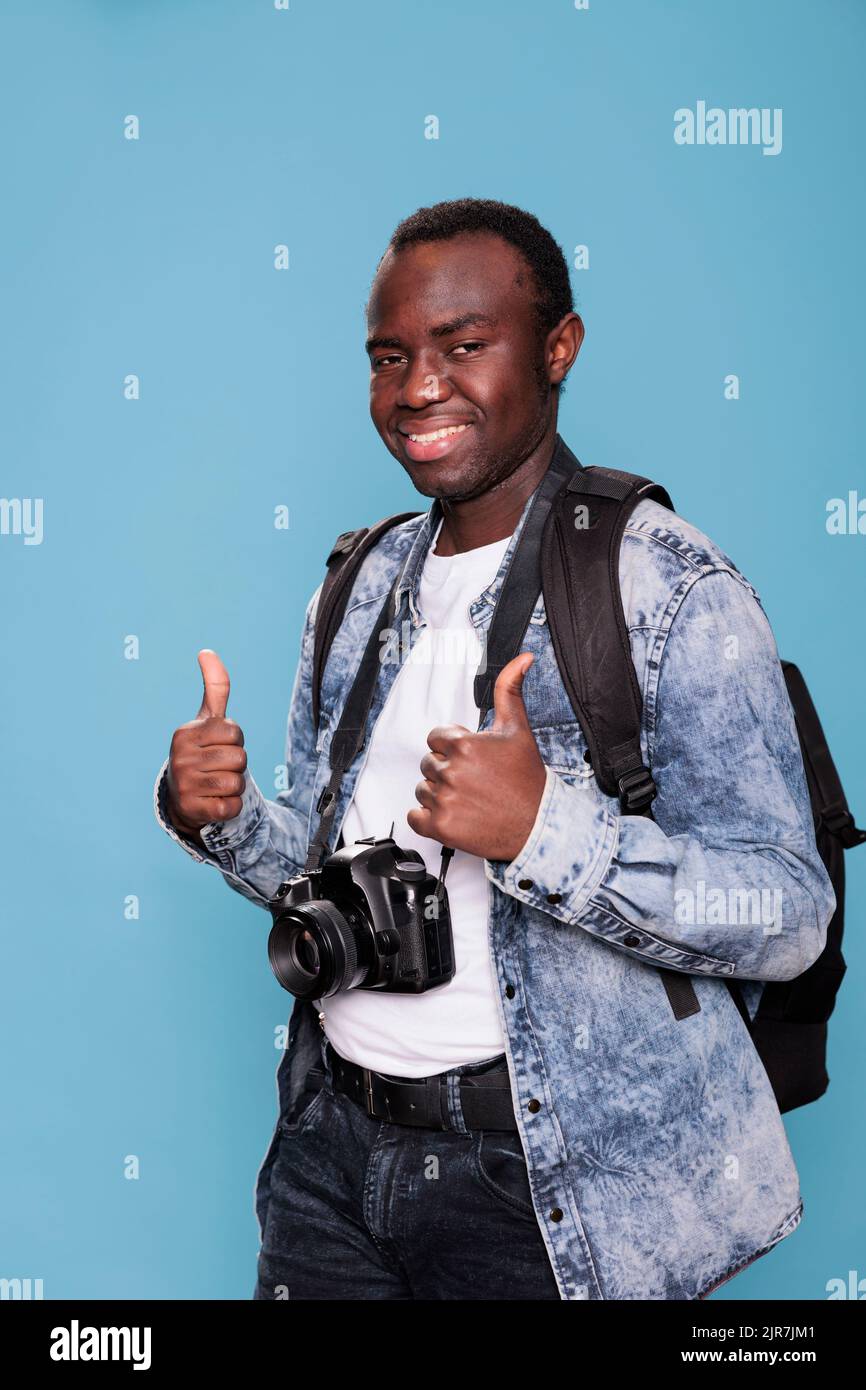 Smiling young man with professional camera giving thumbs up hand symbol ...