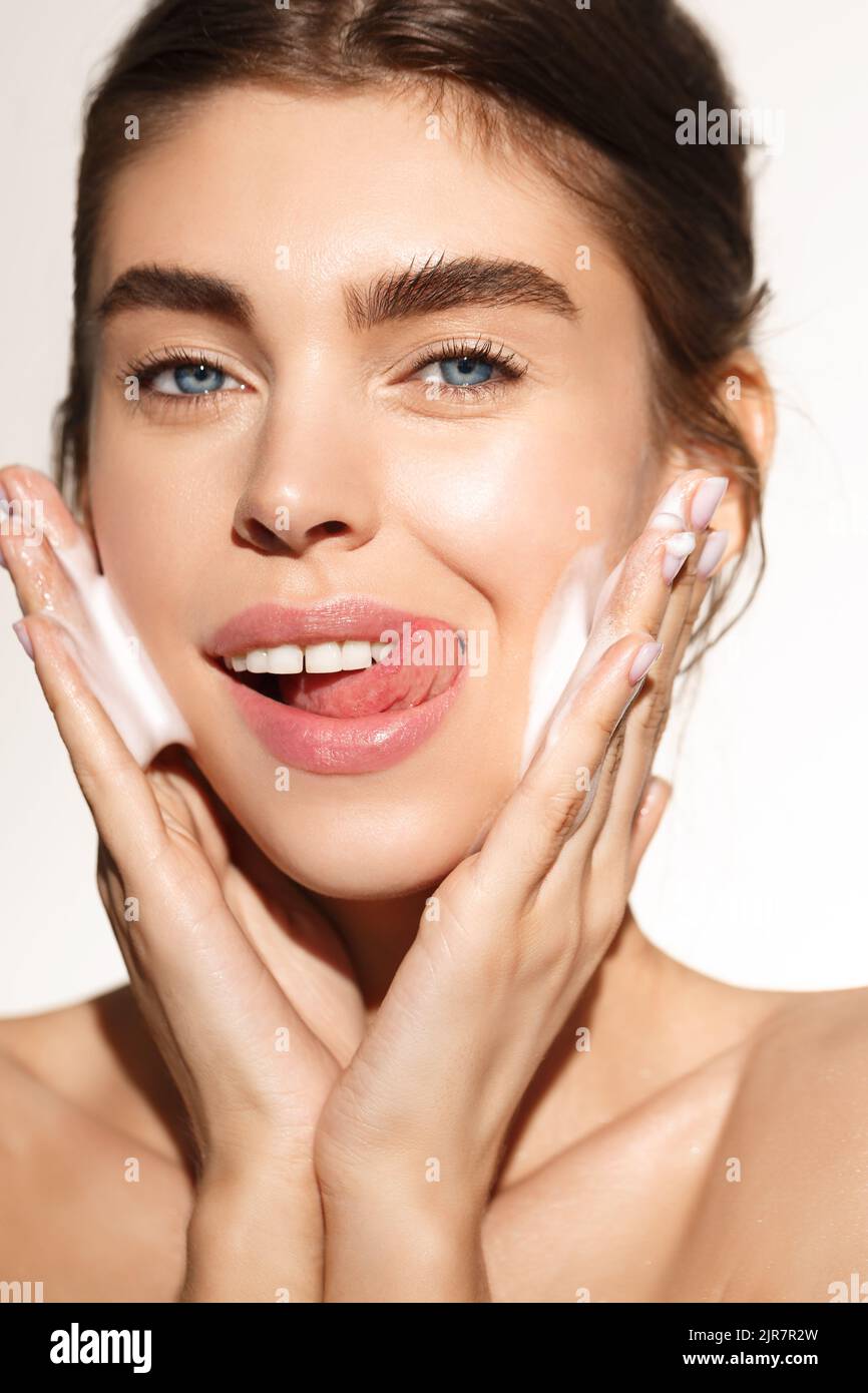 Vertical portrait of young woman washing her face with cleansing foam ...