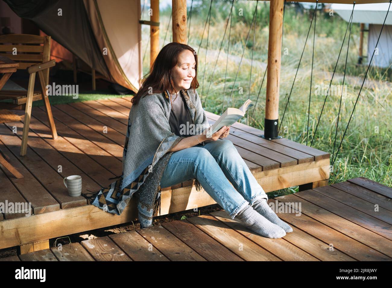 Young brunette woman in poncho reading book and relaxing in glamping in ...