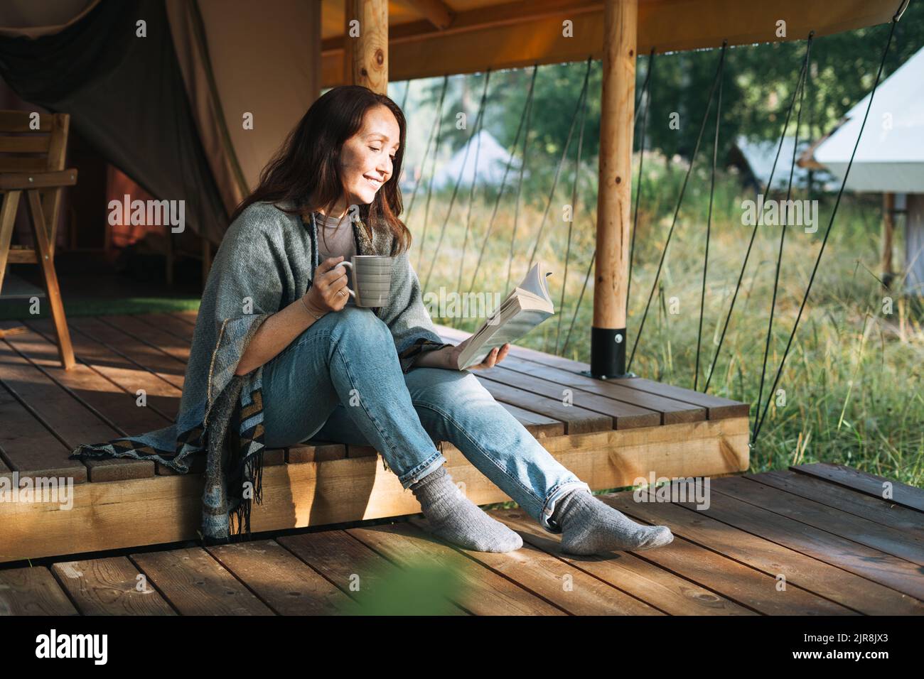 Young brunette woman in poncho reading book and relaxing in glamping in ...