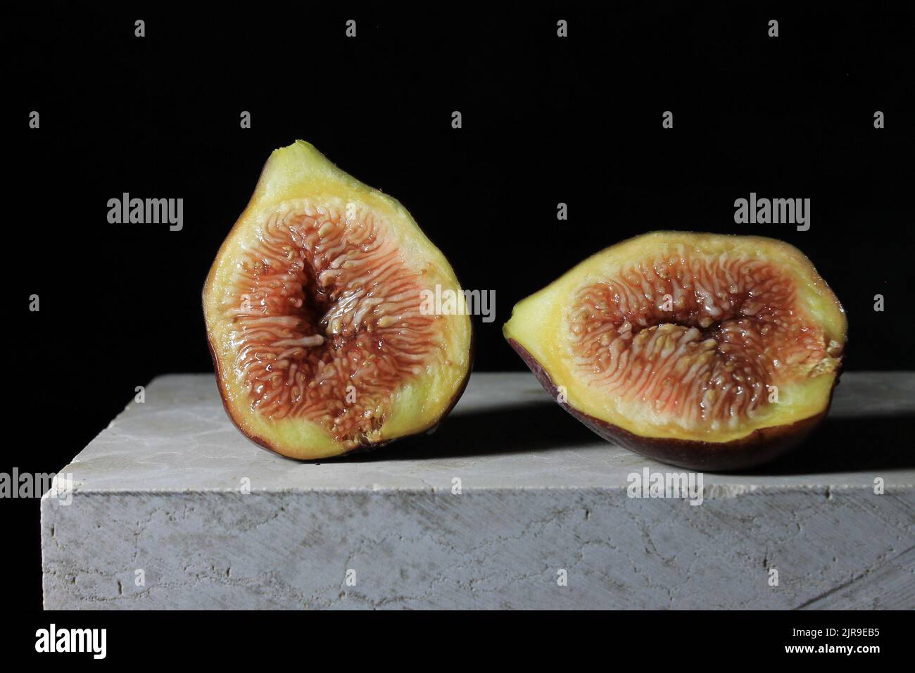 A fresh ripe fig cut in half on white marble against a black background ...
