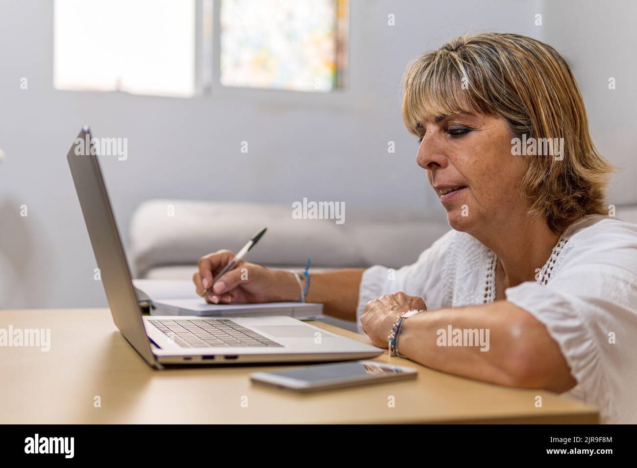 Mature woman sitting and working from home, jotting down notes in her ...