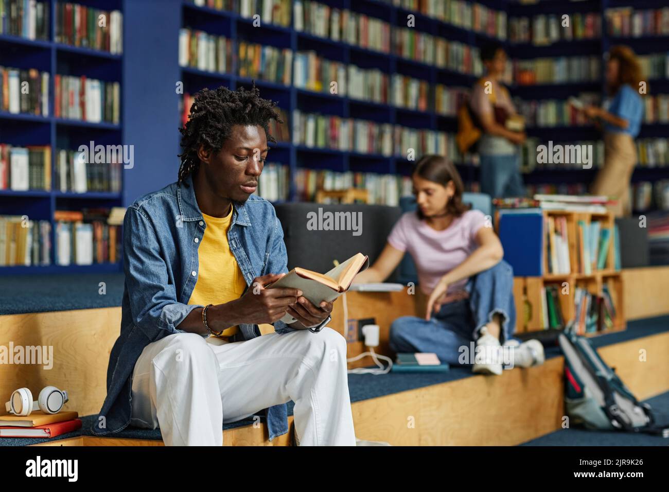 Portrait of young black man reading book in library lounge with ...