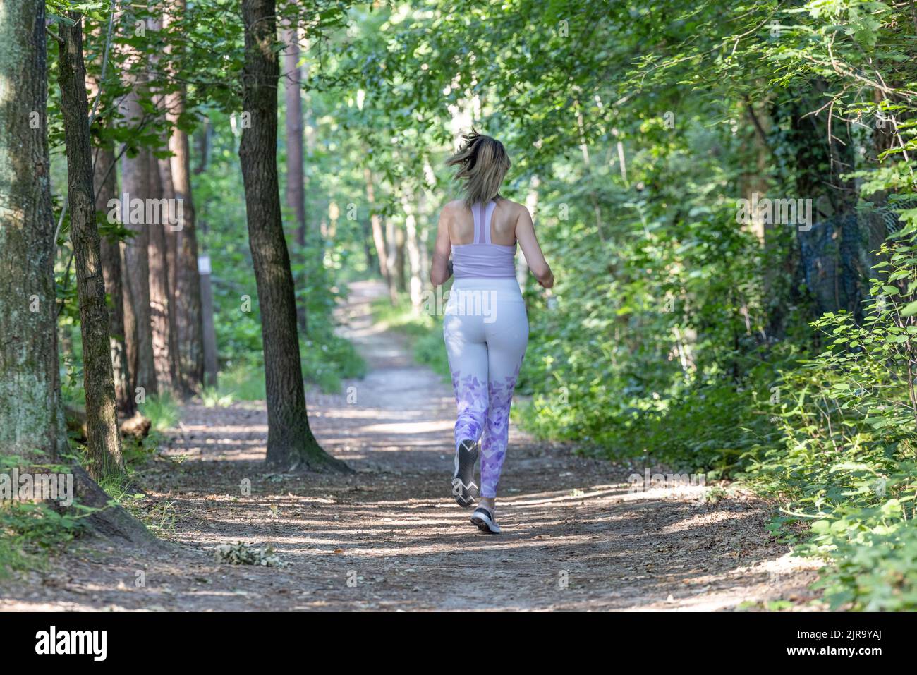Blonde Caucasian young fitness woman running on forest trail in the sun ...