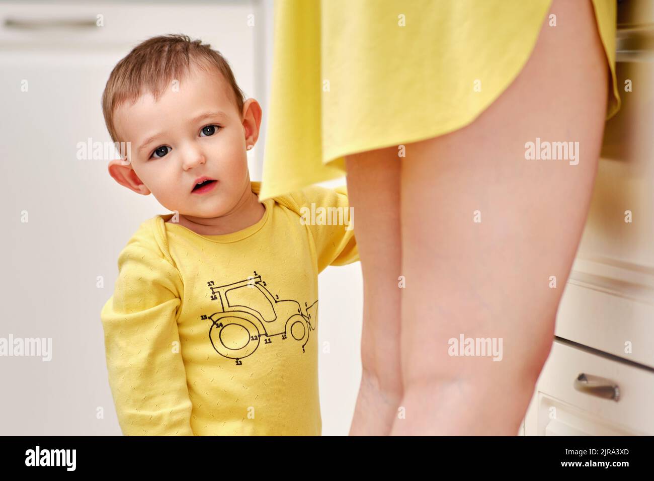 Sad toddler baby boy stands holding his mother legs in the kitchen, kid ...