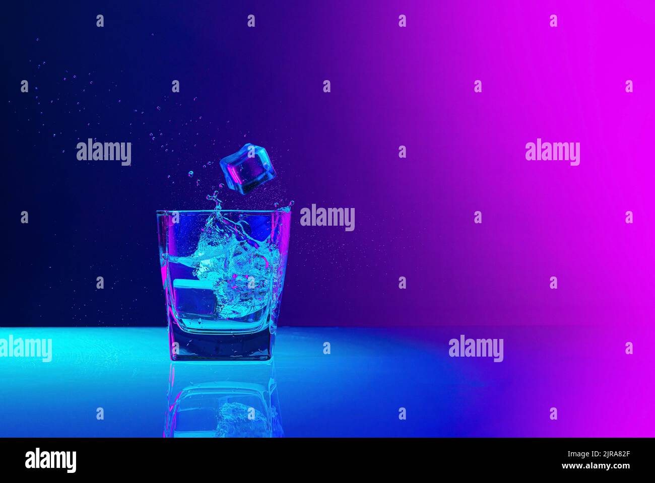 Ice cube falls into a transparent glass of water standing on mirror surface over gradient blue ...