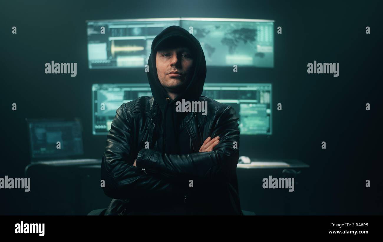 Male Cybercriminal In Hood Looking At Camera In Front Of Table With Data Hacking Computers In