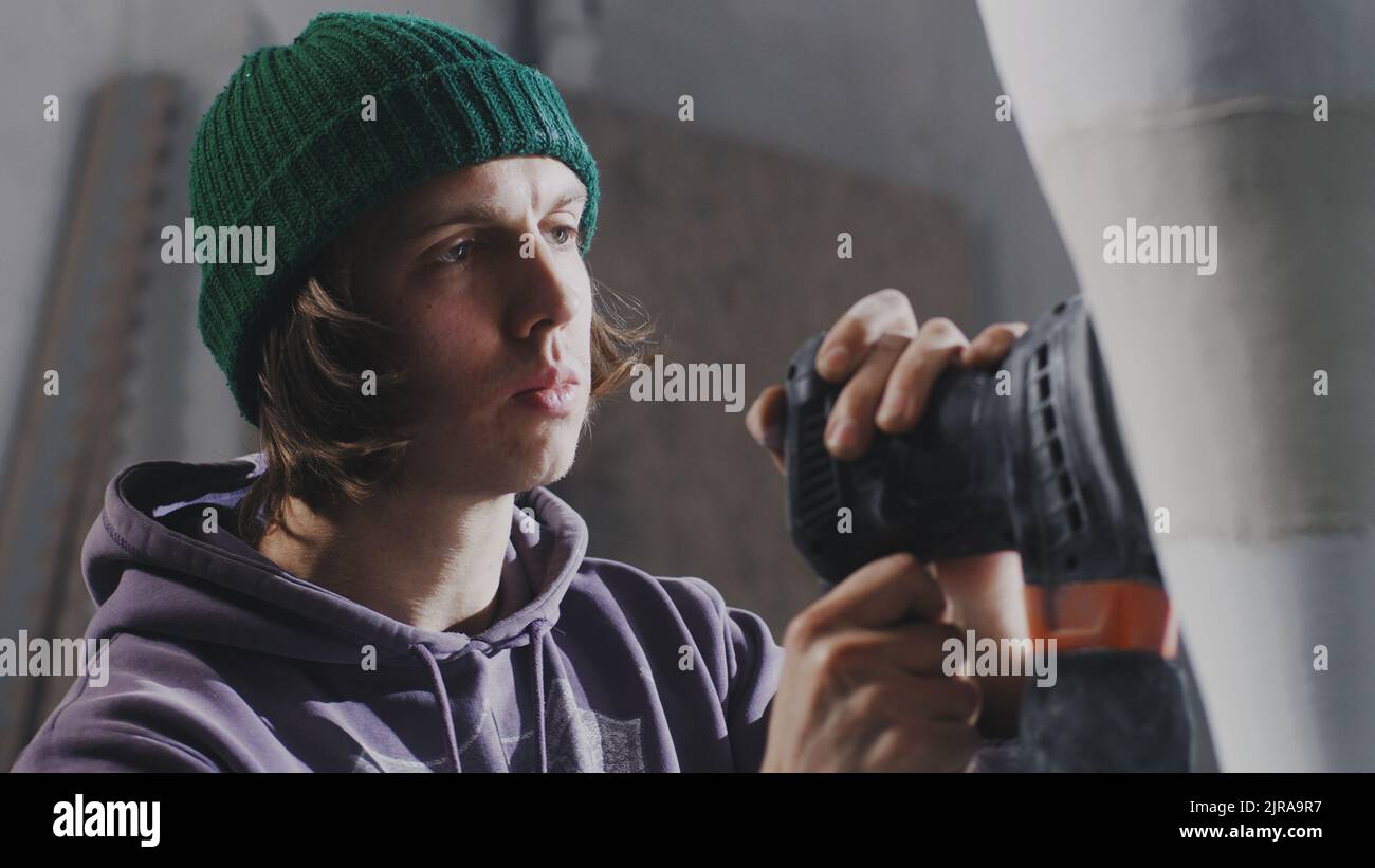 Low angle of young male artisan in hoodie and hat using sander to ...