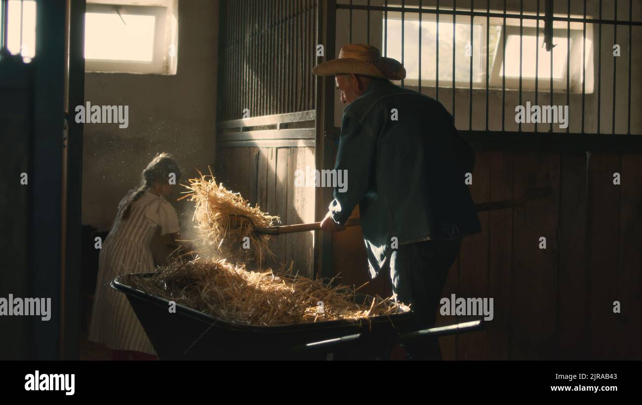 Elderly man in casual clothes and hat using pitchfork to throw straw ...