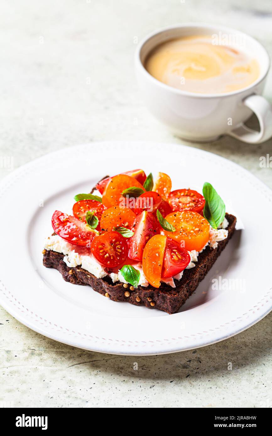 Rye toast with cream cheese, tomatoes and basil on white plate and cup ...