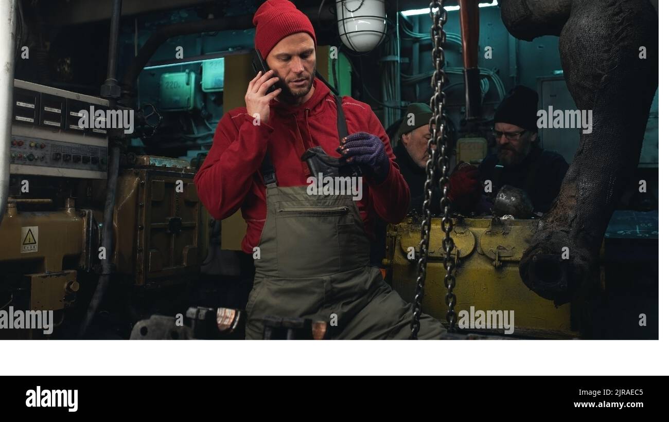 Zoom in view of adult man examining metal part and talking on mobile phone near colleagues ...