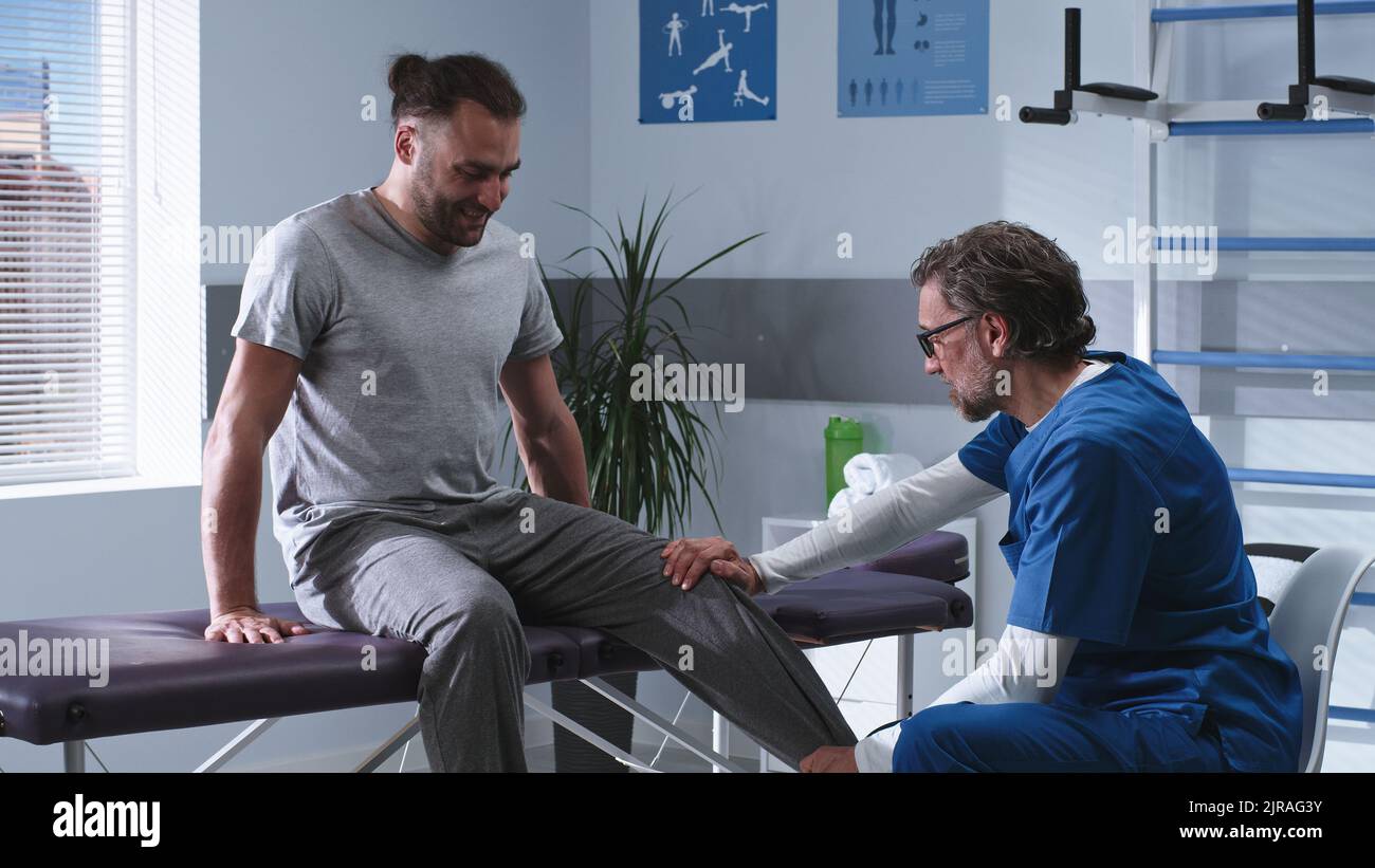 Middle aged male doctor stretching injured leg of strained man sitting ...