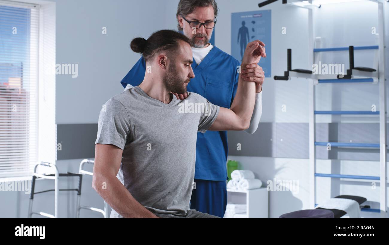 Mature doctor massaging and stretching arm of adult male patient during ...