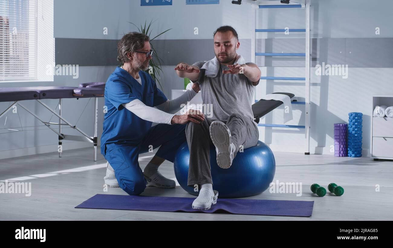 Mature male doctor supporting adult man balancing on fit ball during ...