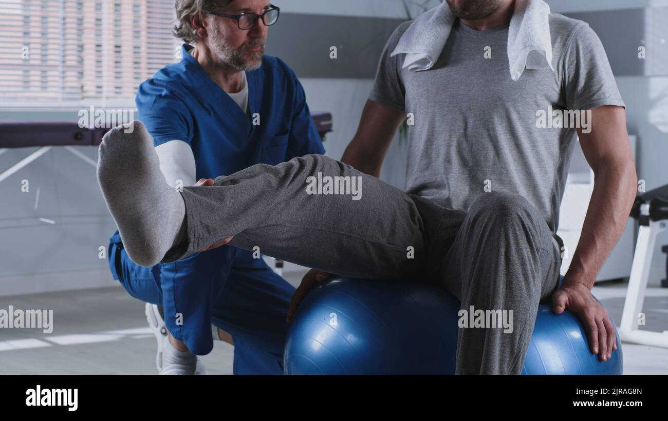 Middle aged doctor stretching leg of man sitting on fit ball during ...