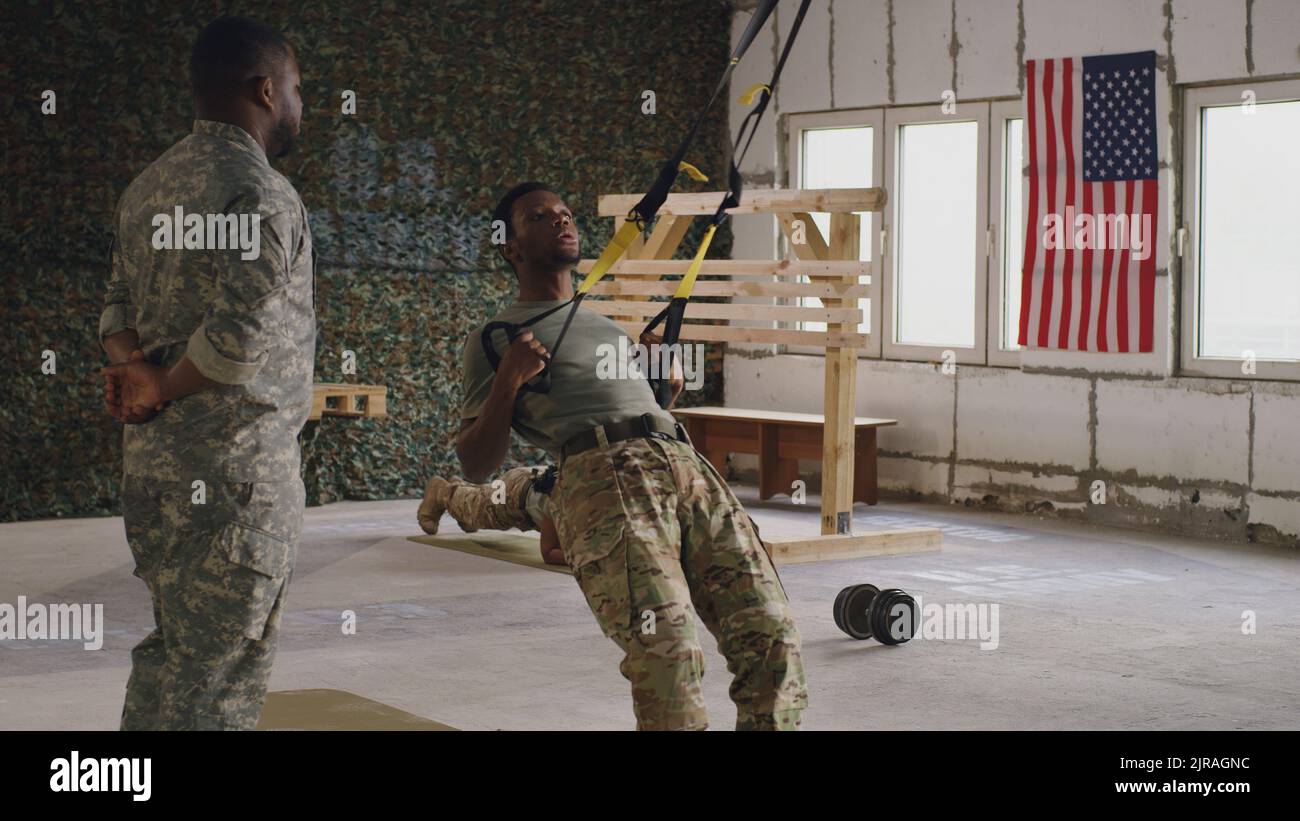 Black instructor talking to male soldier exercising with TRX ropes ...