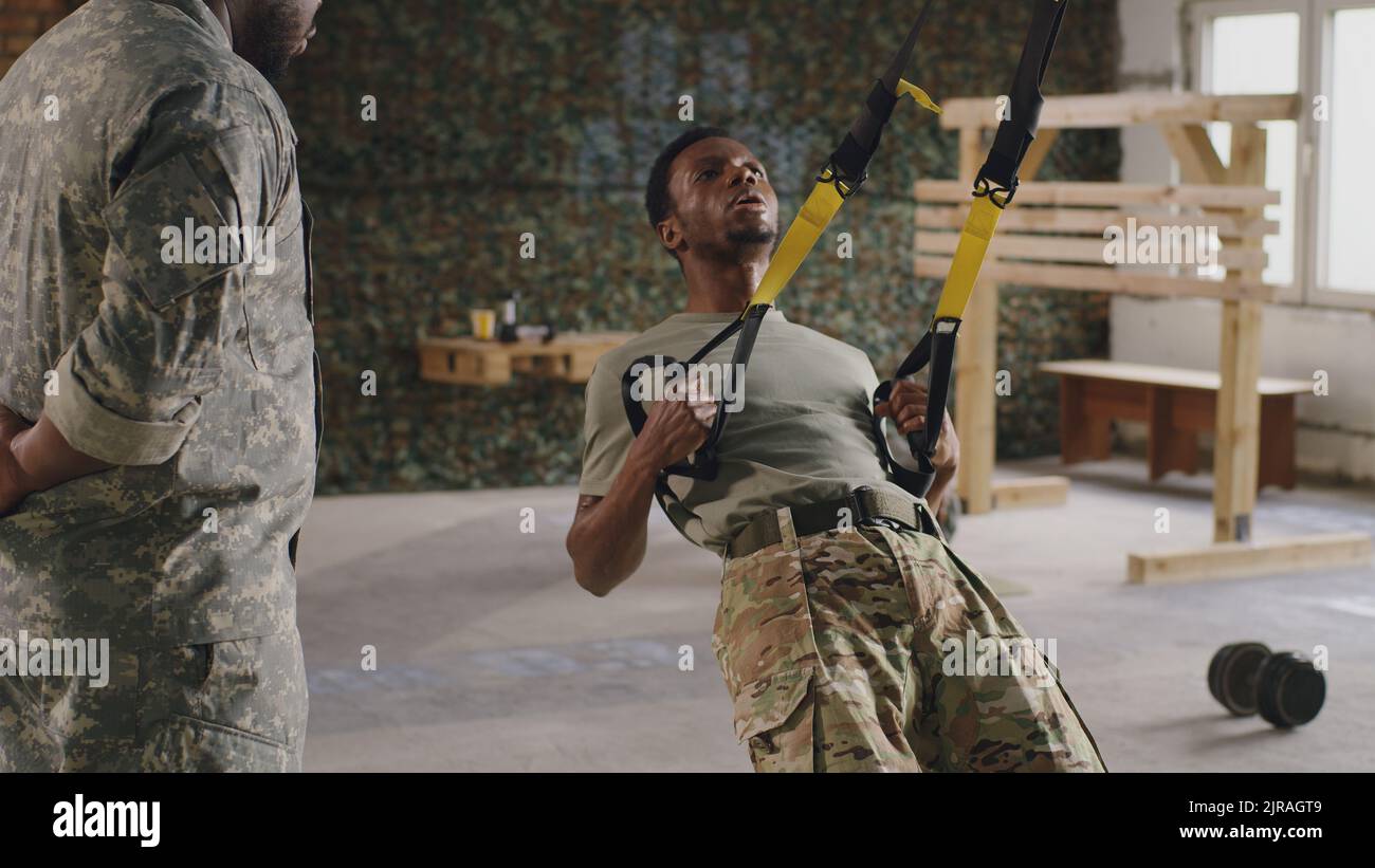 Black instructor talking to male soldier exercising with TRX ropes ...