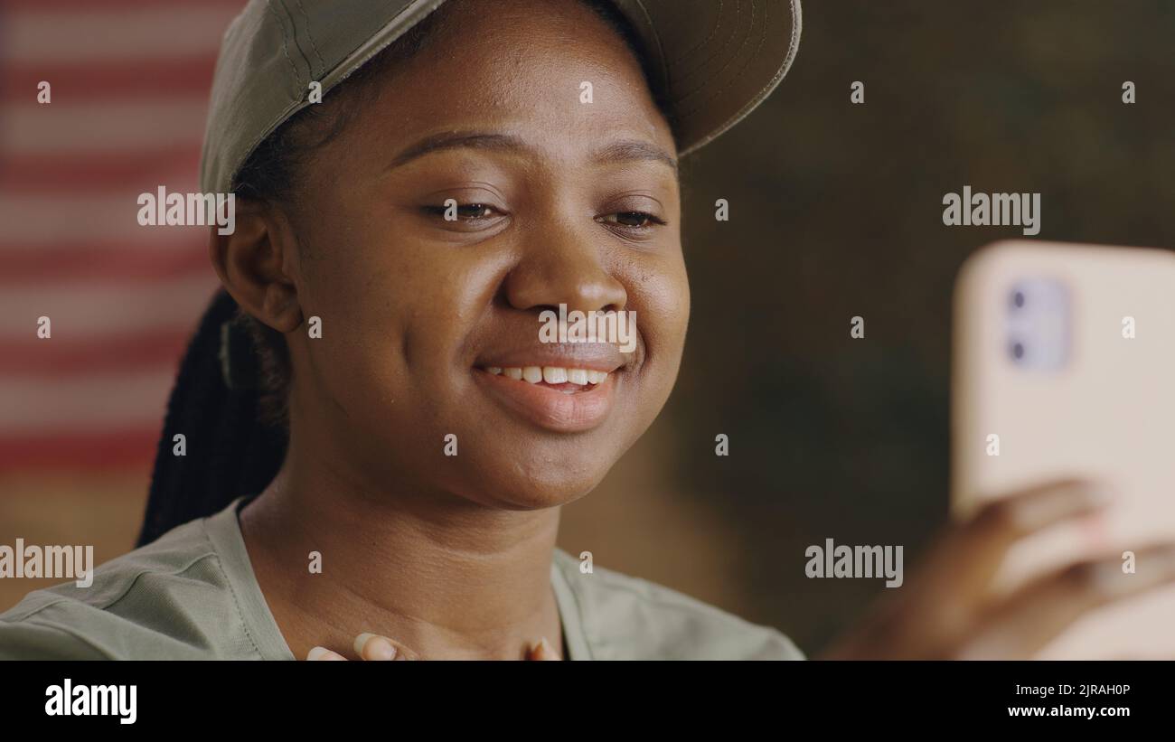 Delighted black military woman waving hand and sharing news with online friend while making ...