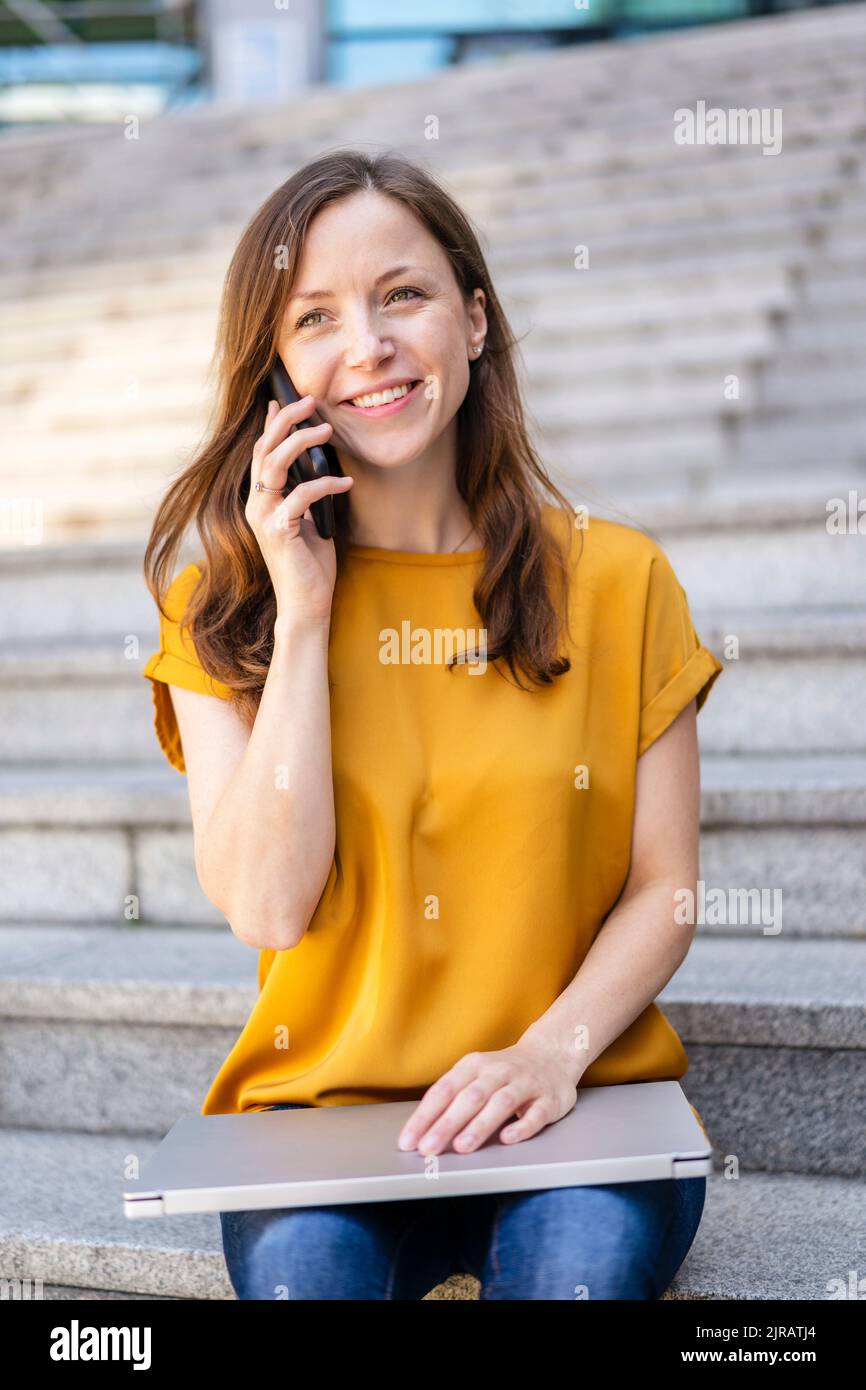 Smiling freelancer with laptop talking through smart phone sitting on ...