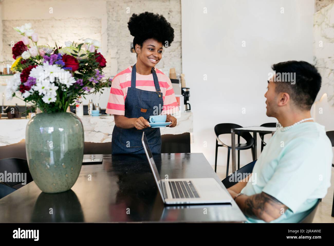 Smiling waitress serving coffee to customer sitting with laptop in cafe ...