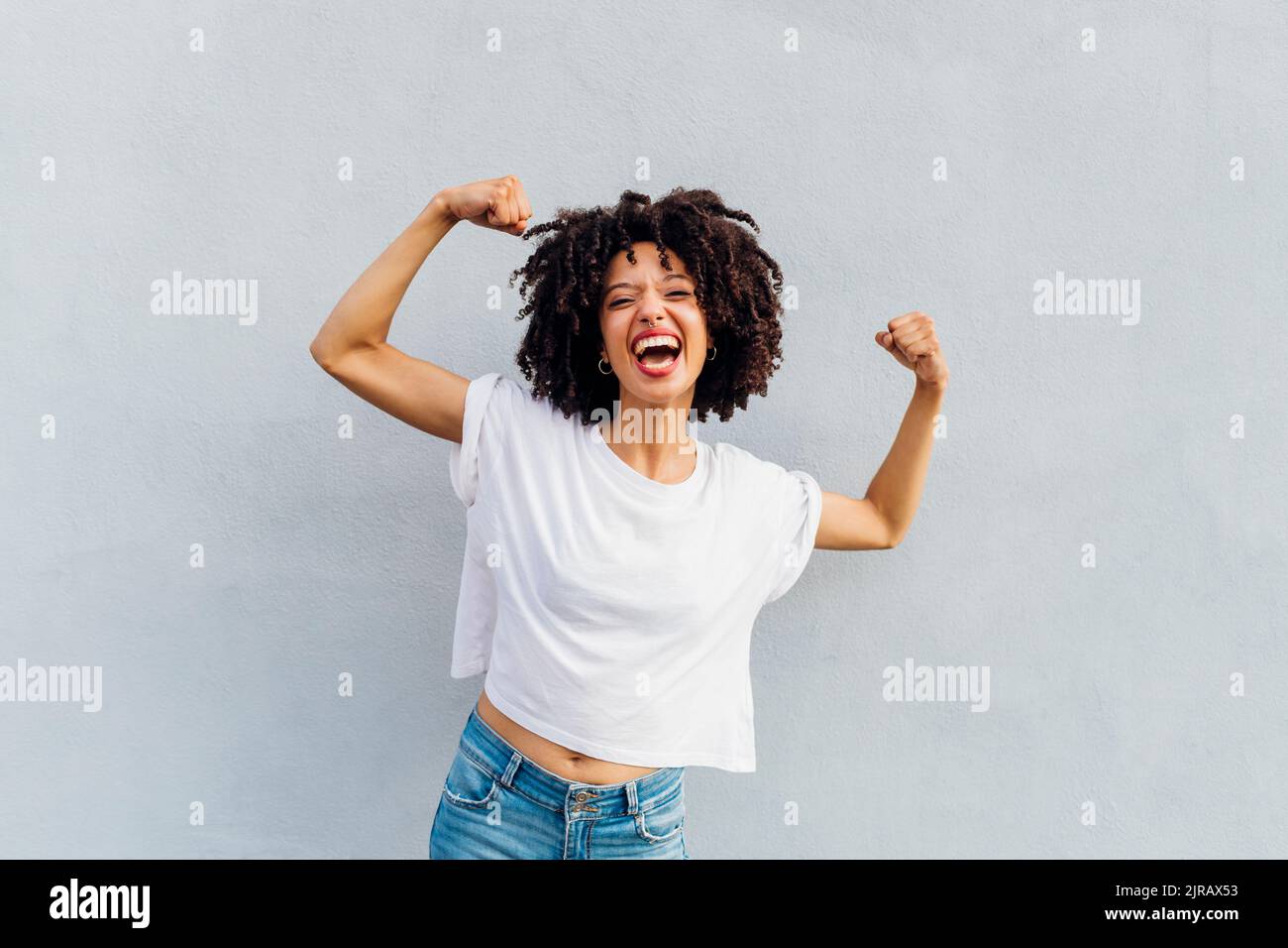 Cheerful young woman flexing muscles in front of white wall Stock Photo ...
