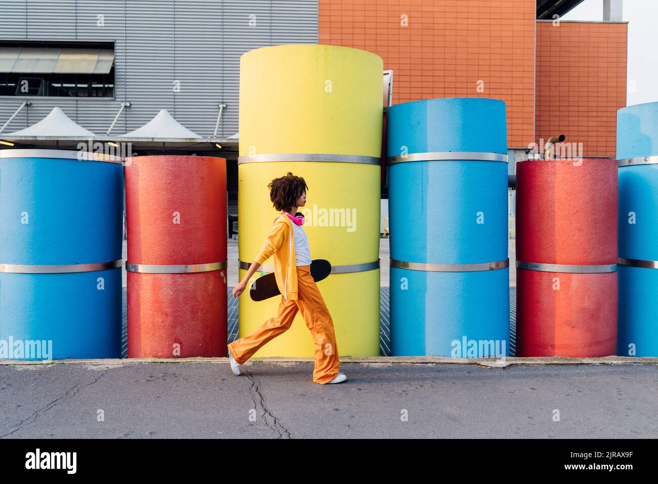 Young woman holding skateboard walking by multi colored pipes Stock ...