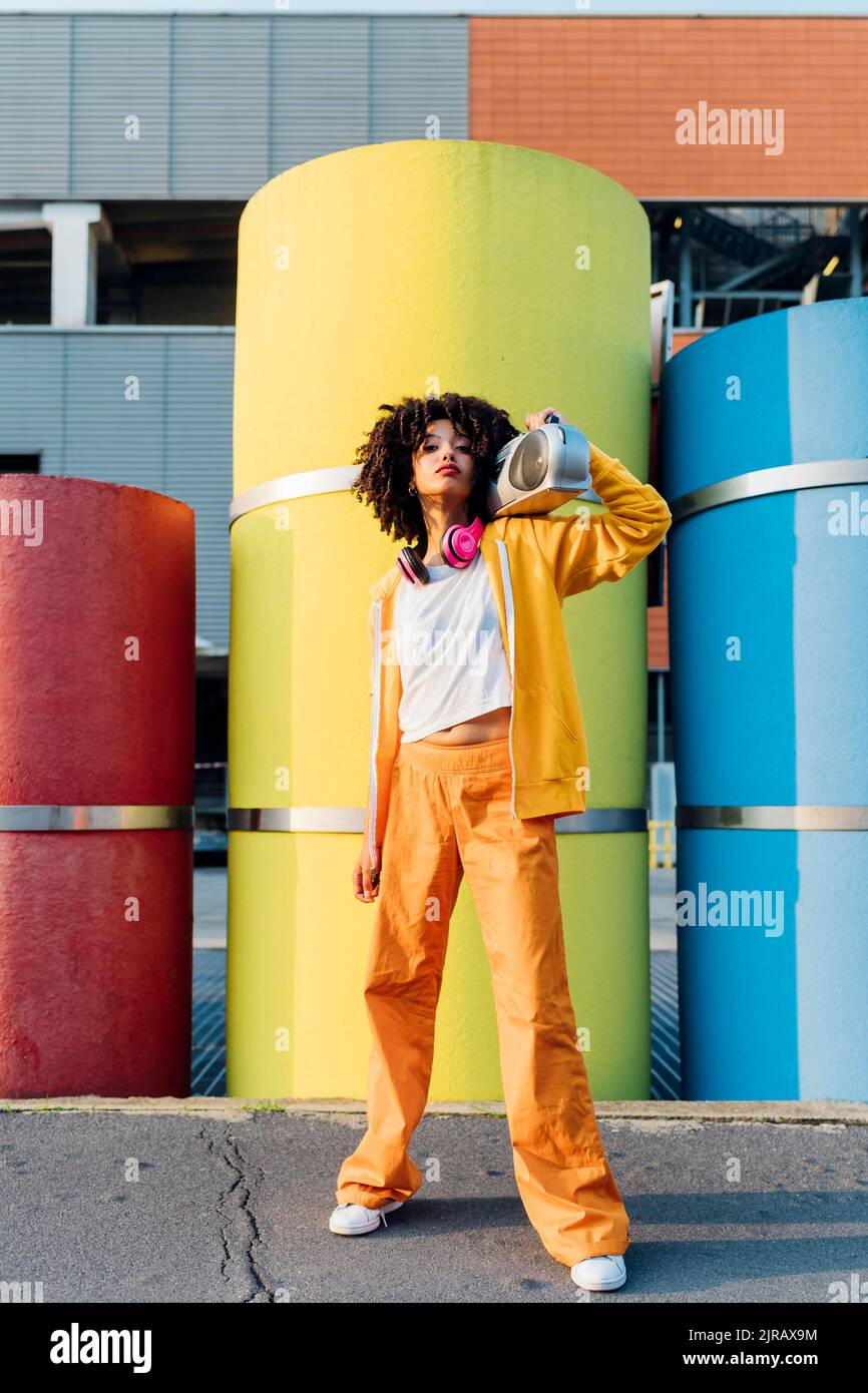 Young woman carrying boom box on shoulder in front of colorful pipes ...