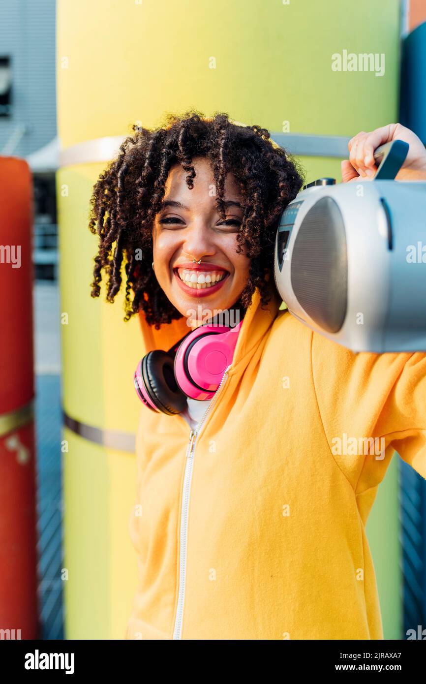 Happy woman carrying boom box on shoulder standing in front of yellow ...