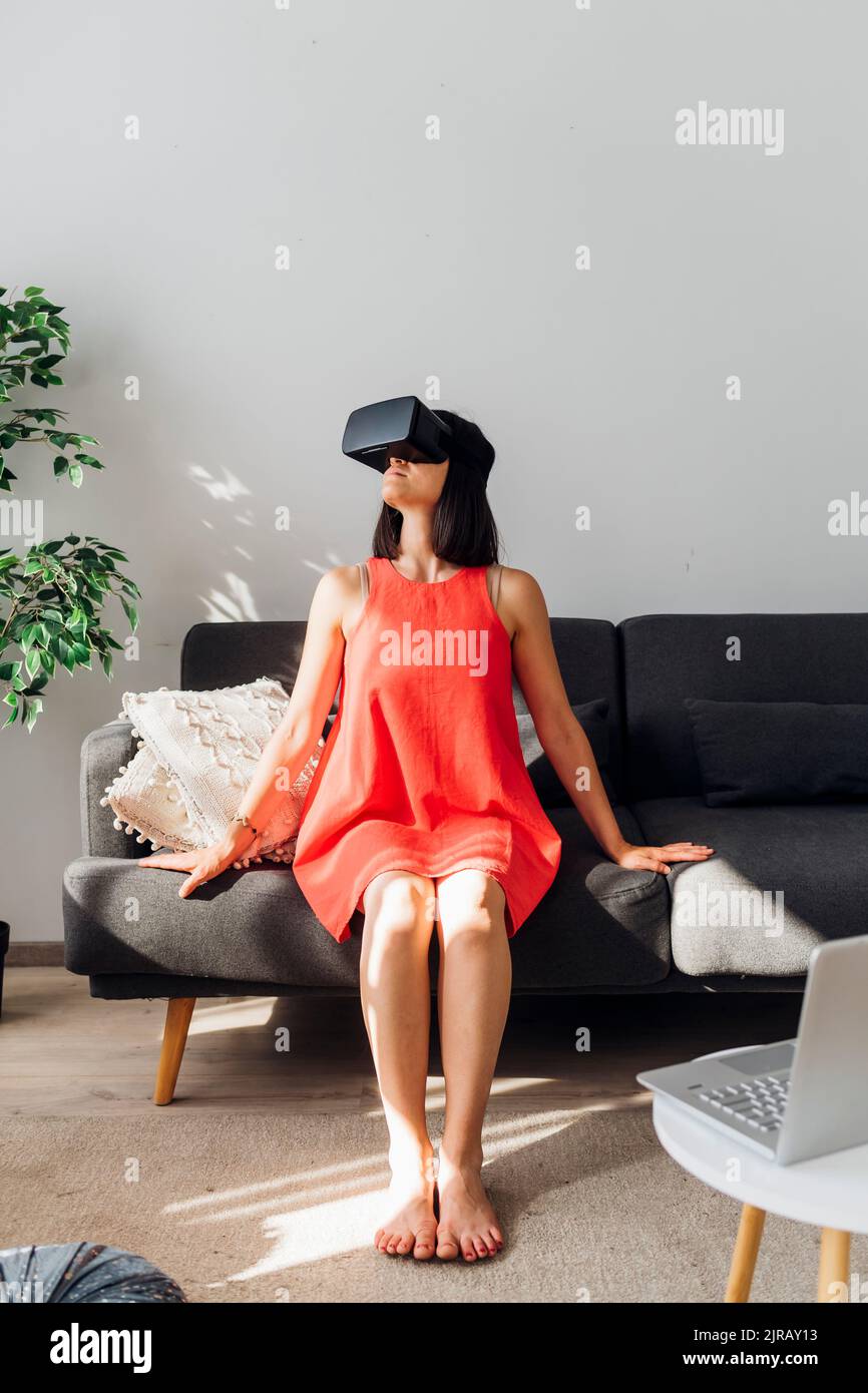 Woman wearing virtual reality simulator sitting on couch in living room ...