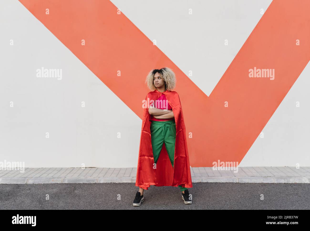 Young heroine wearing cape standing with arms crossed in front of wall ...