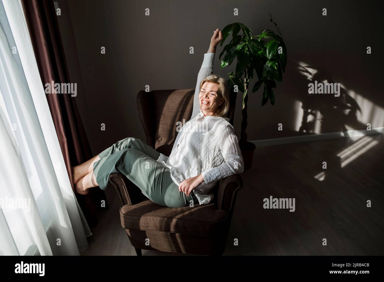 Cheerful mature woman sitting with arms raised in chair at home Stock ...