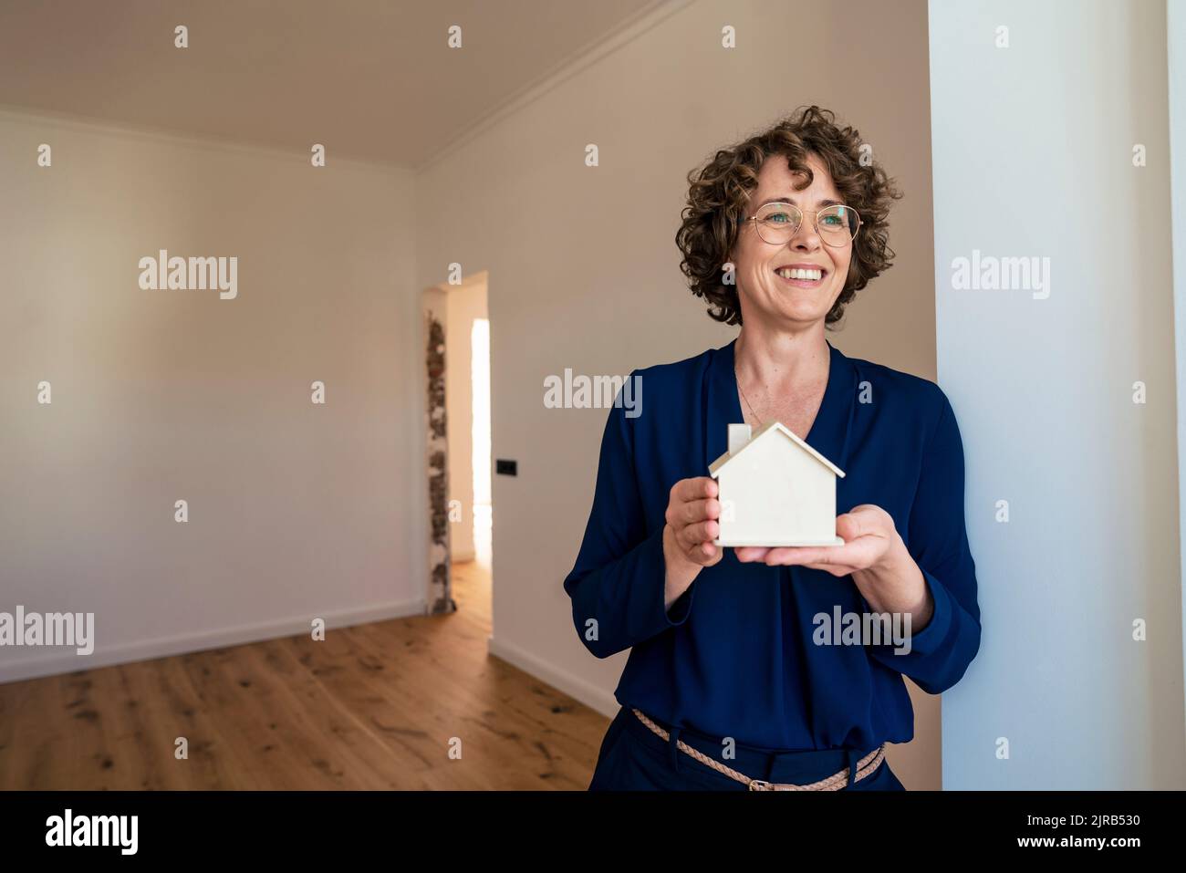 Smiling real estate agent with model house leaning on wall in apartment ...