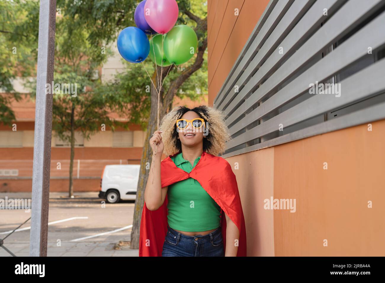 Smiling woman wearing cape standing with multi colored balloons by wall ...