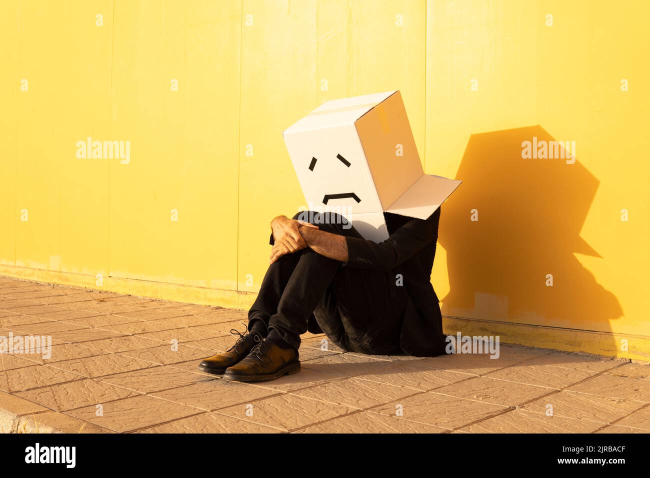 Man wearing box with sad face sitting on footpath Stock Photo - Alamy