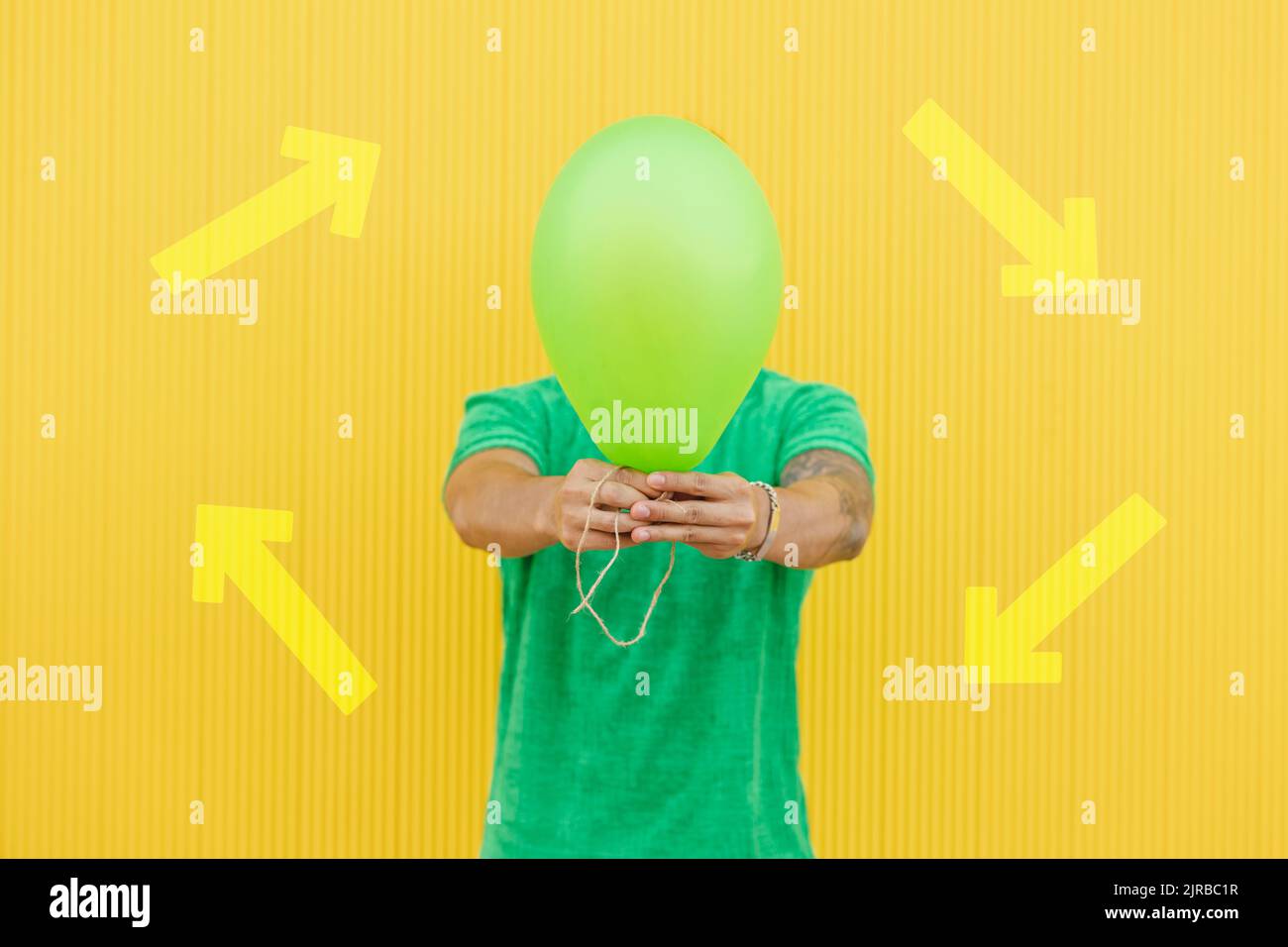 Young man covering face with green balloon in front of arrow symbols on ...
