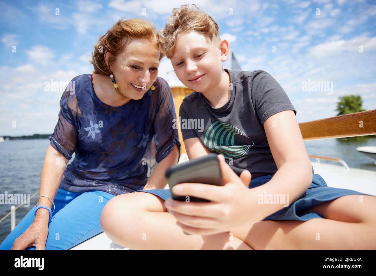 Grandson sharing mobile phone with grandmother sitting on boat deck ...