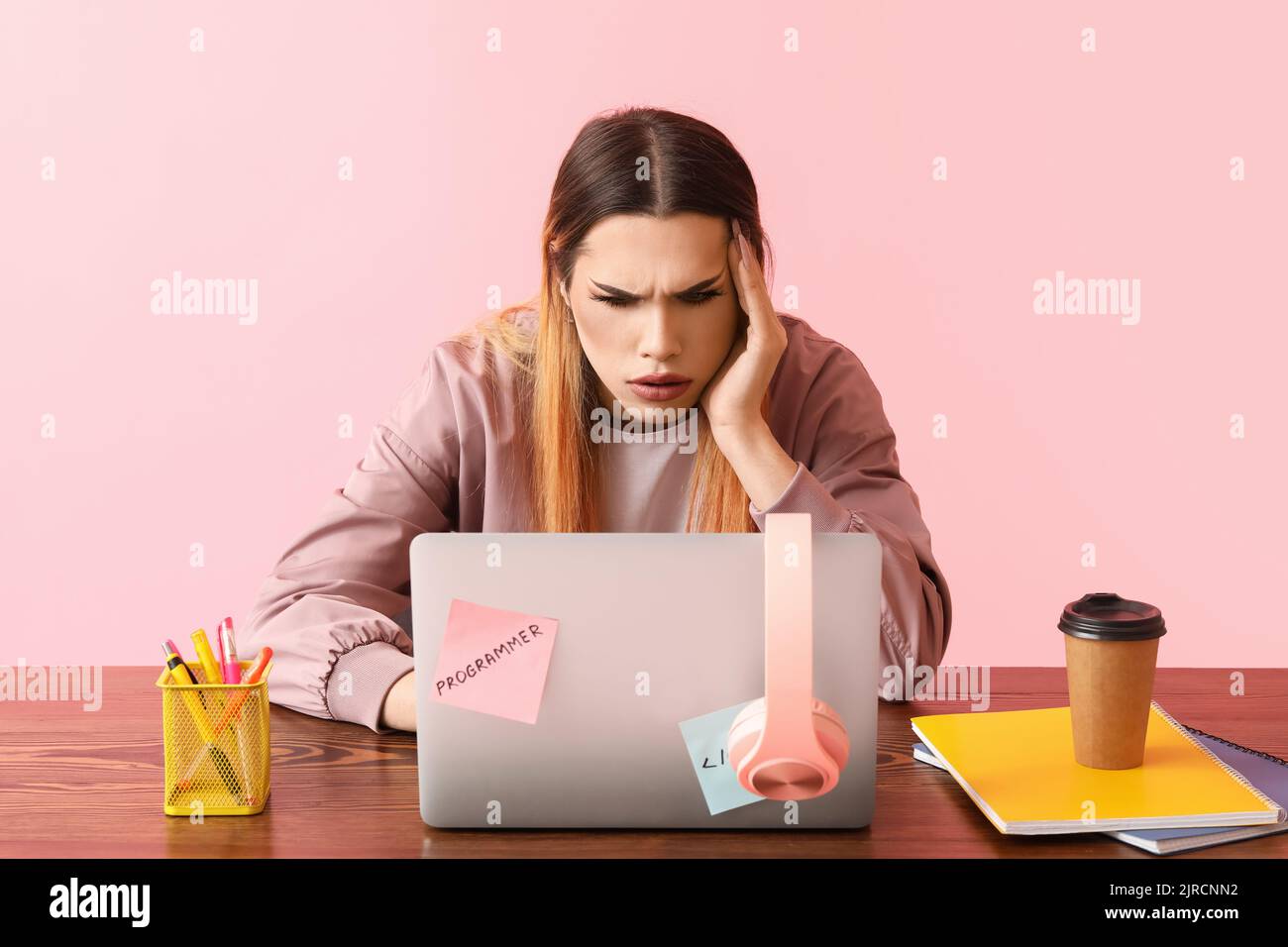 Displeased transgender programmer with laptop at table on pink ...