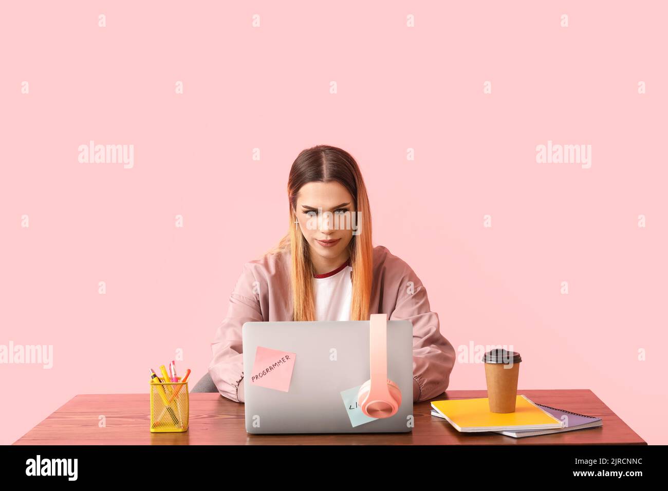 Transgender programmer with laptop at table on pink background Stock ...