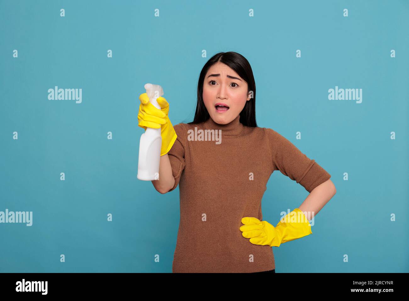 Angrily surprised young asian housewife holding spray in yellow glove ...