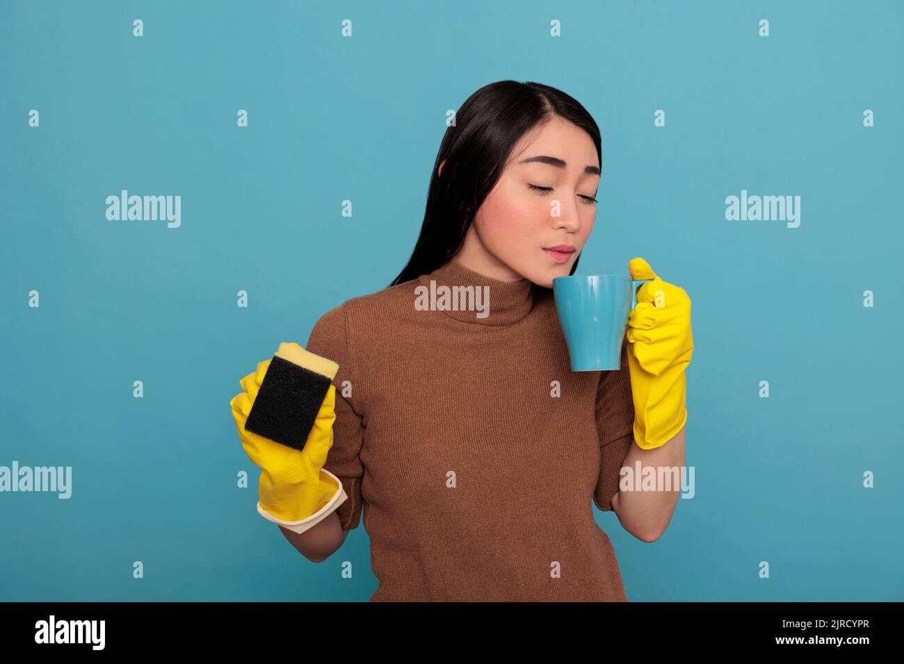 Glad happy asian young house maid wearing yellow gloves taking coffee ...