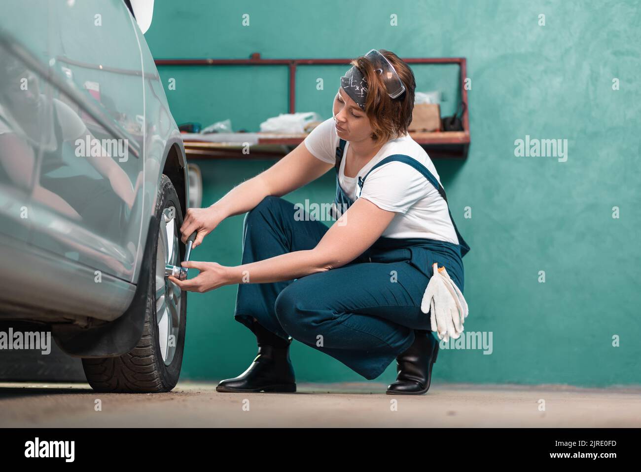 Garage. Young woman mechanic in blue coveralls squatting and repair ...